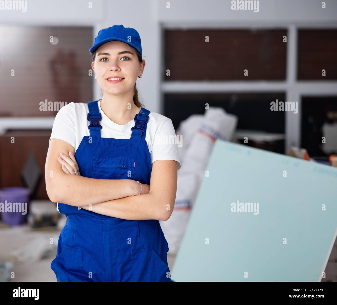 Portrait of positive builder woman in blue overalls next to stepladder ...