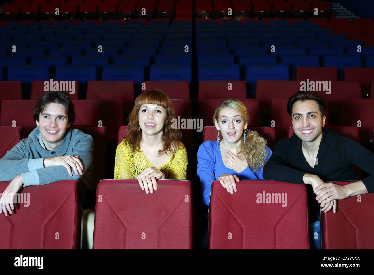 Four young smiling friends sit on seats in cinema theater and see movie ...