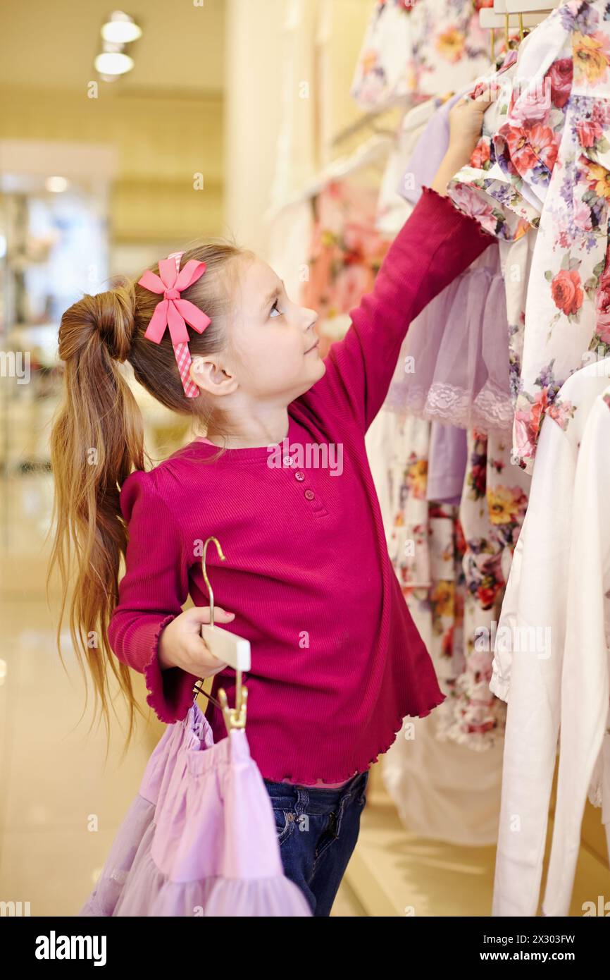 Little girl takes off hangers with dresses from stand in clothing store ...