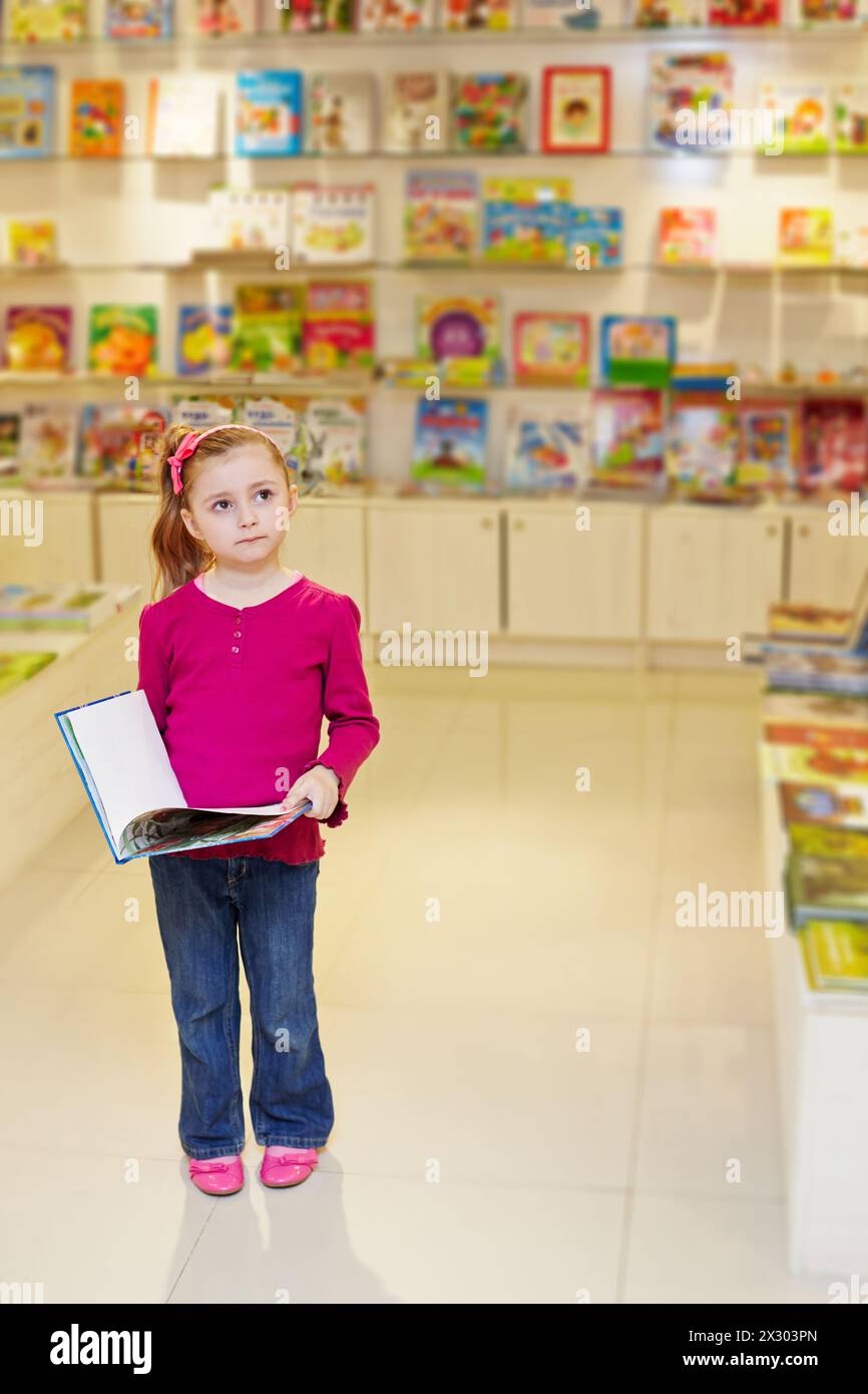 Little girl stands looking thoughtful holding open book in book ...