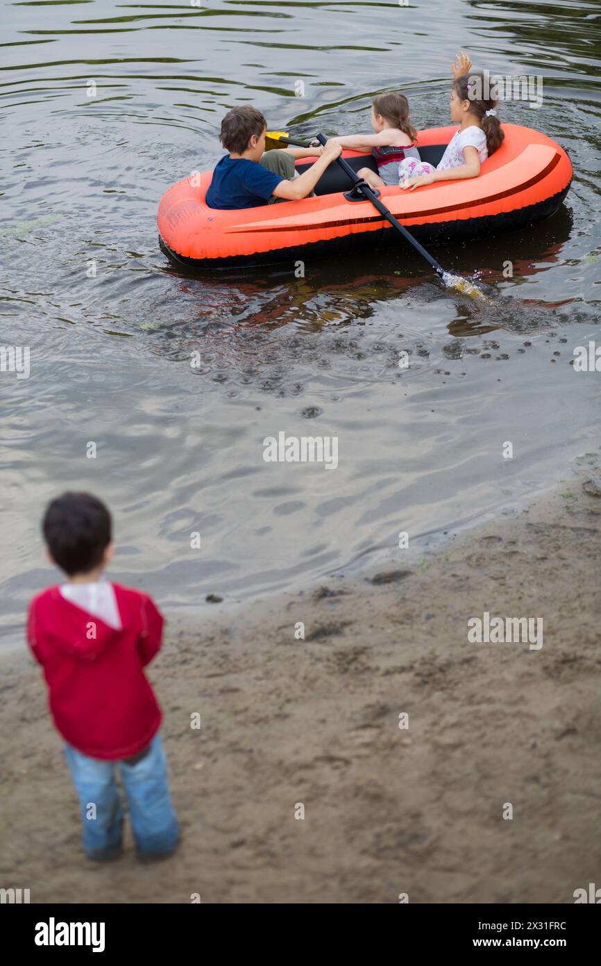Three children floating in a rubber boat on the pond, one boy standing ...