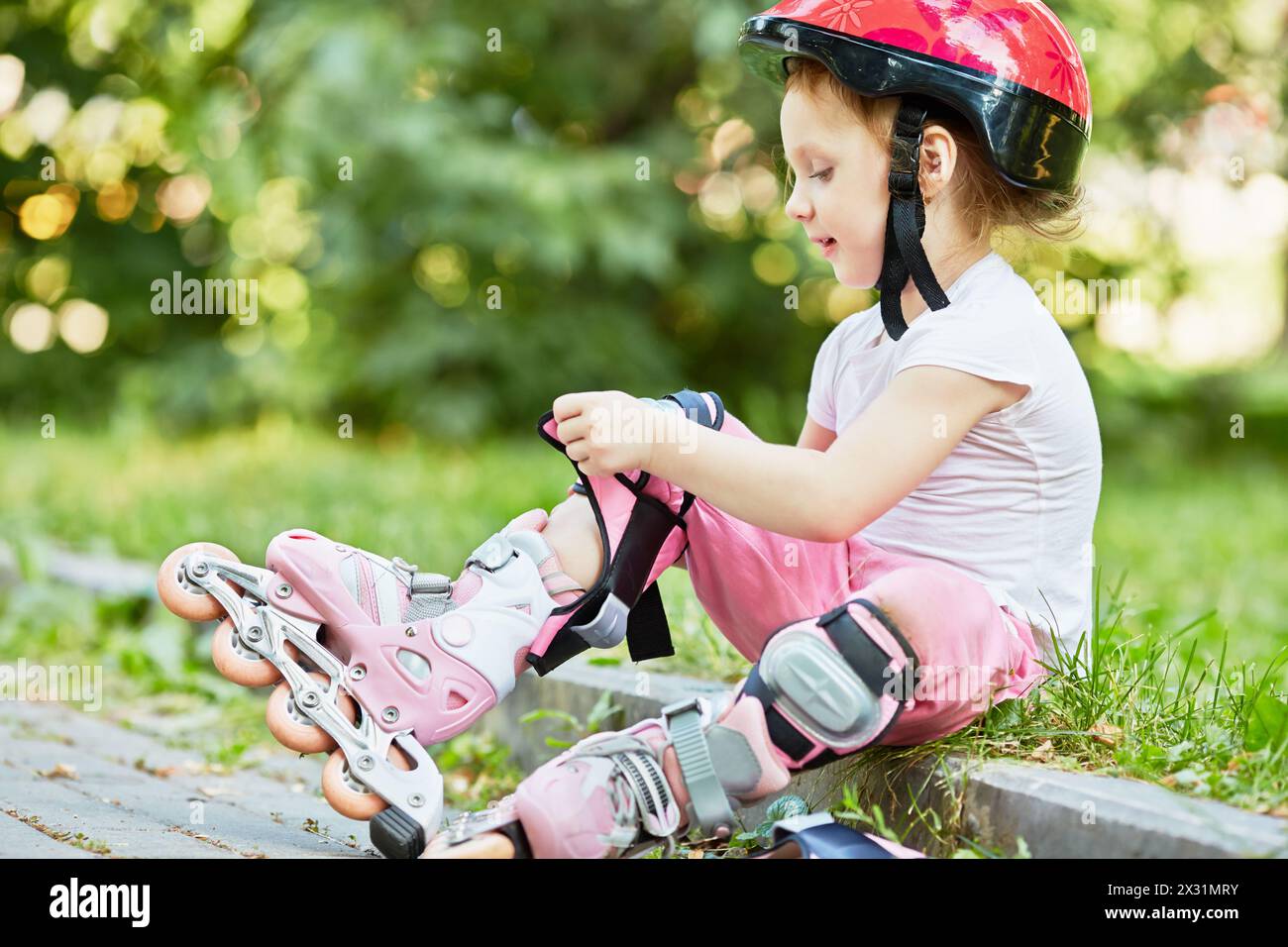 Little girl in protective equipment and rollers sits on curb of walkway ...