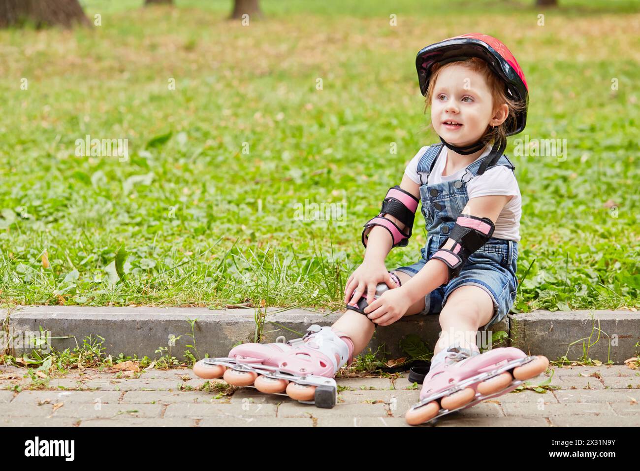 Little girl in protective equipment and rollers sits resting on curb of ...