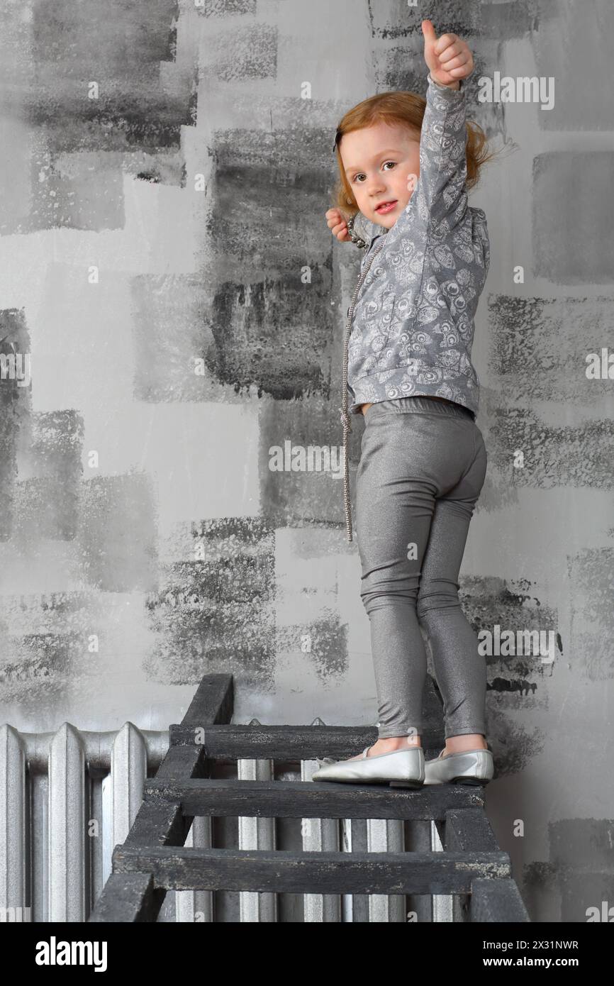 A little girl with hand raised standing on ladder at gray wall Stock ...