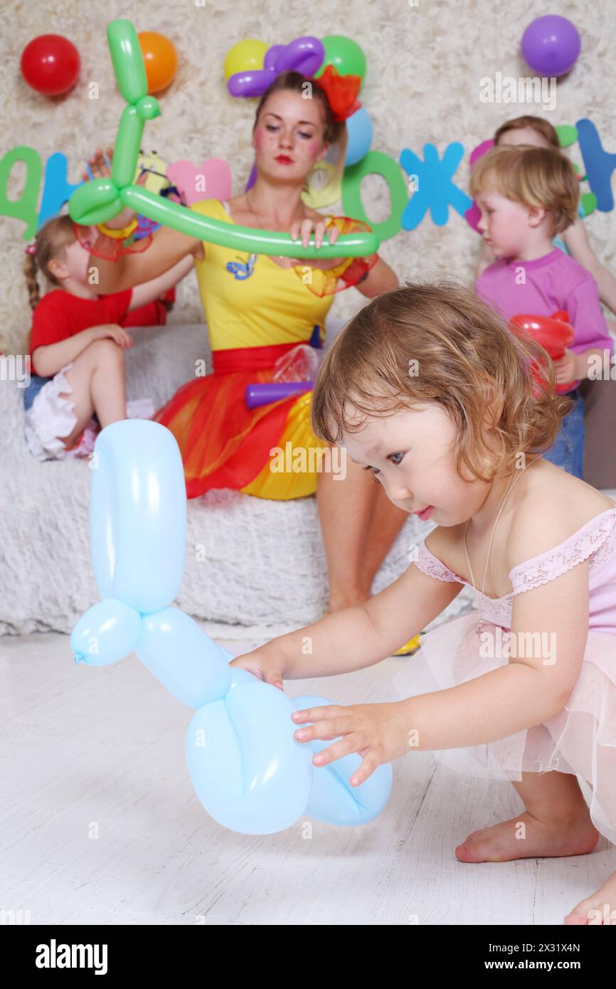 Four happy kids and facilitator make pets of long balloons at children party. Focus on girl with ...