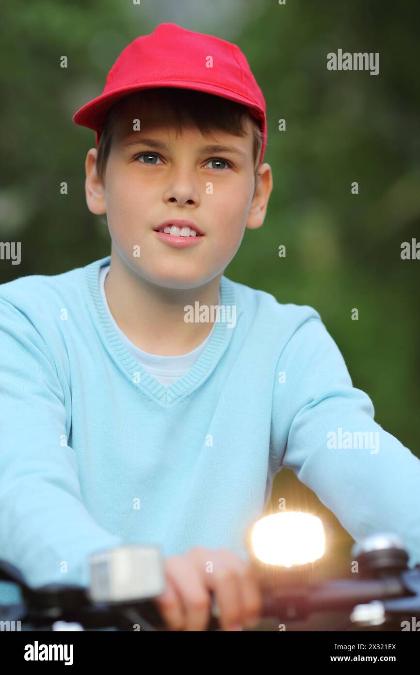 Happy boy in red cap rides bicycle and looks away in park at summer day ...