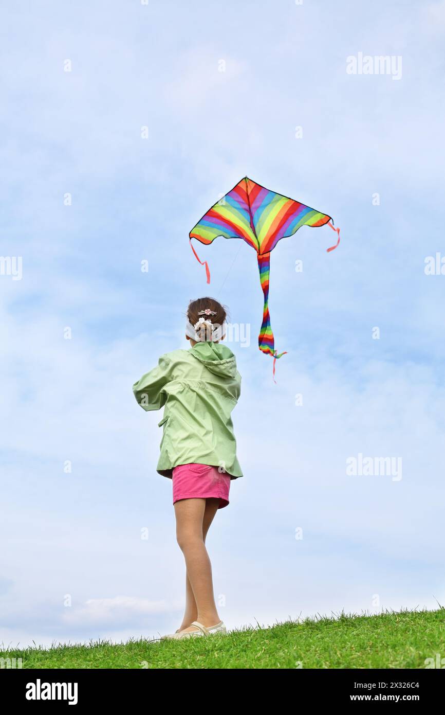 Back view of girl in pink skirt and green jacket launches colorful kite ...