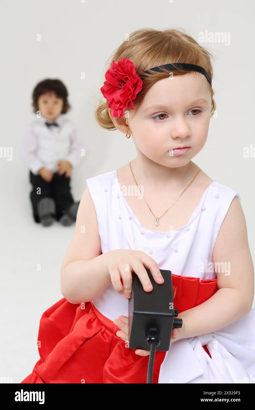 Little girl holds black control console and boy sits behind she on grey ...