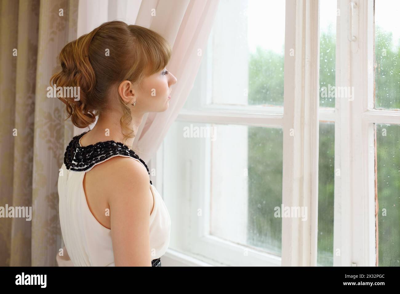 Beautiful sad girl in white dress looks in window in light room Stock ...