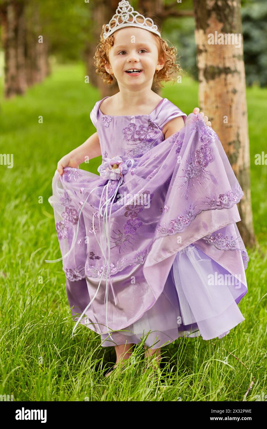 Little girl in beautiful violet gown and crown on head stands on grassy ...