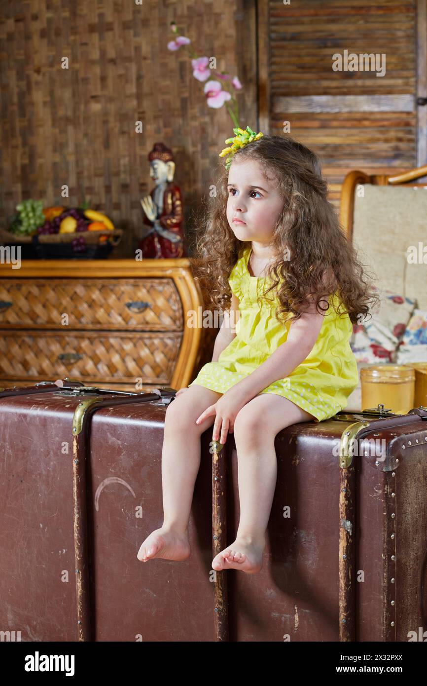 Little bearfooted girl in yellow dress sits on old big ragged suitcase ...