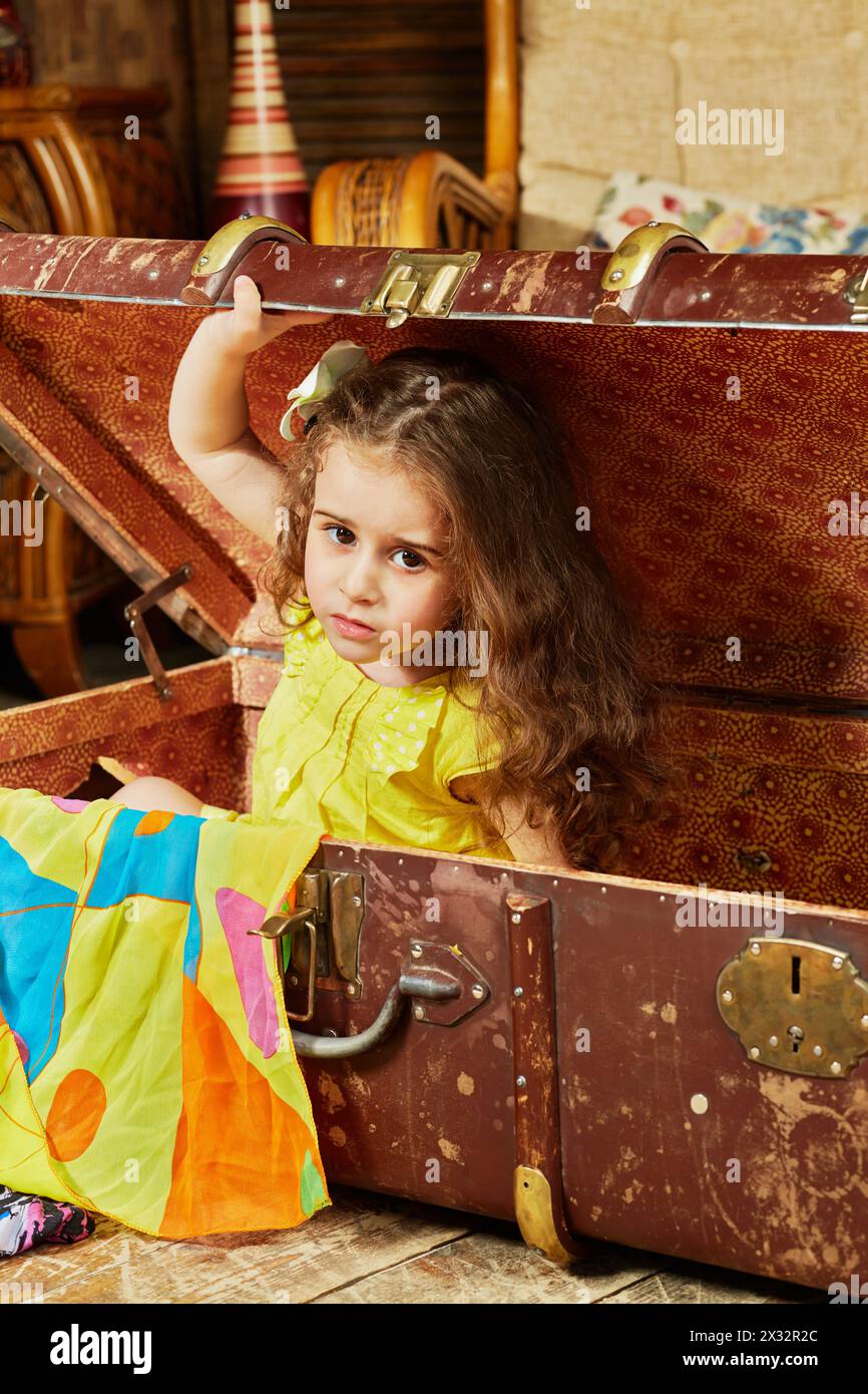 Little girl in yellow dress gets out from old big ragged fiber suitcase ...