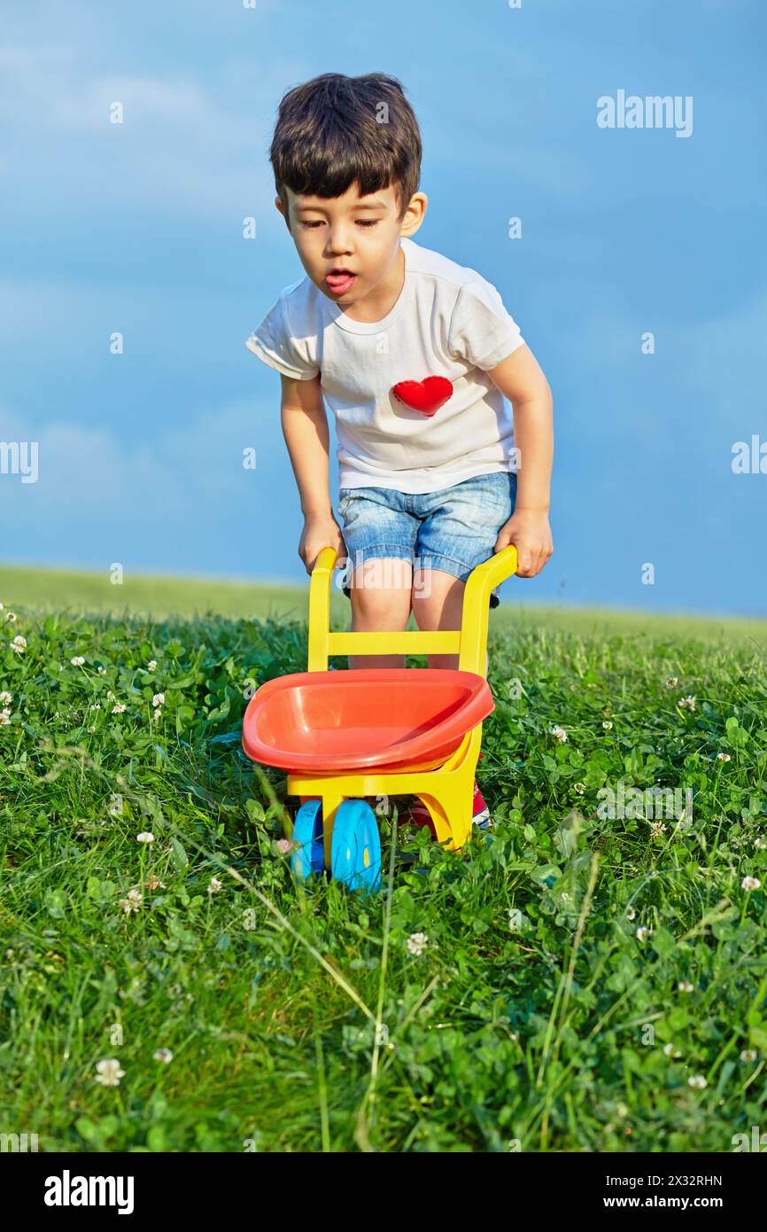 Little boy carries plastic wheelbarrow on grassy slope, big meadow and ...
