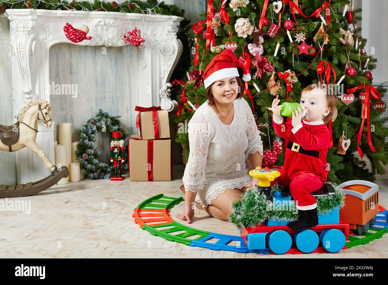 Mother in santa cap sits on floor under decorated Christmas tree near ...