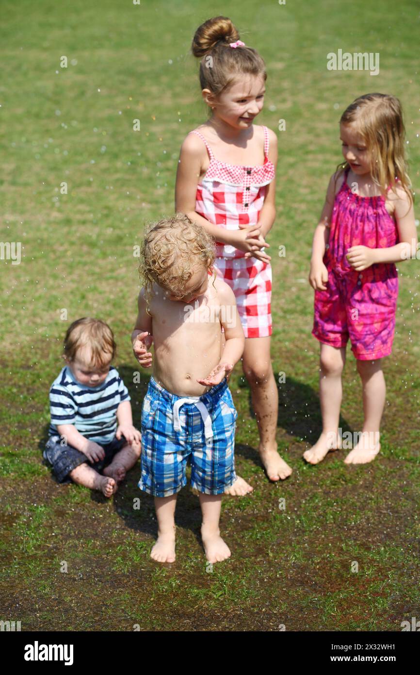 Four wet little funny children stand on grass among spray at summer day ...