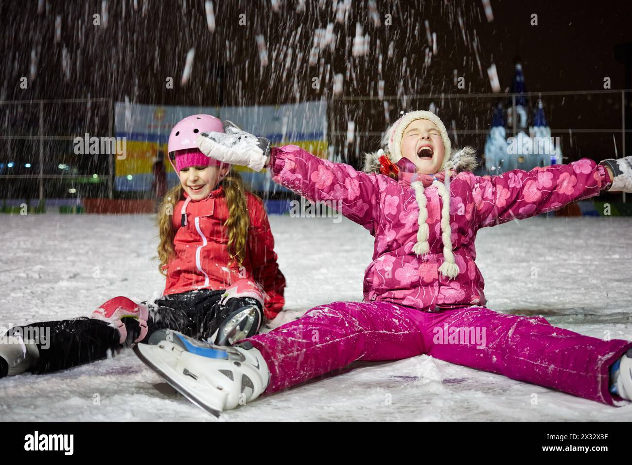 Two little girls sit on skating rink ice in evening under falling snow ...