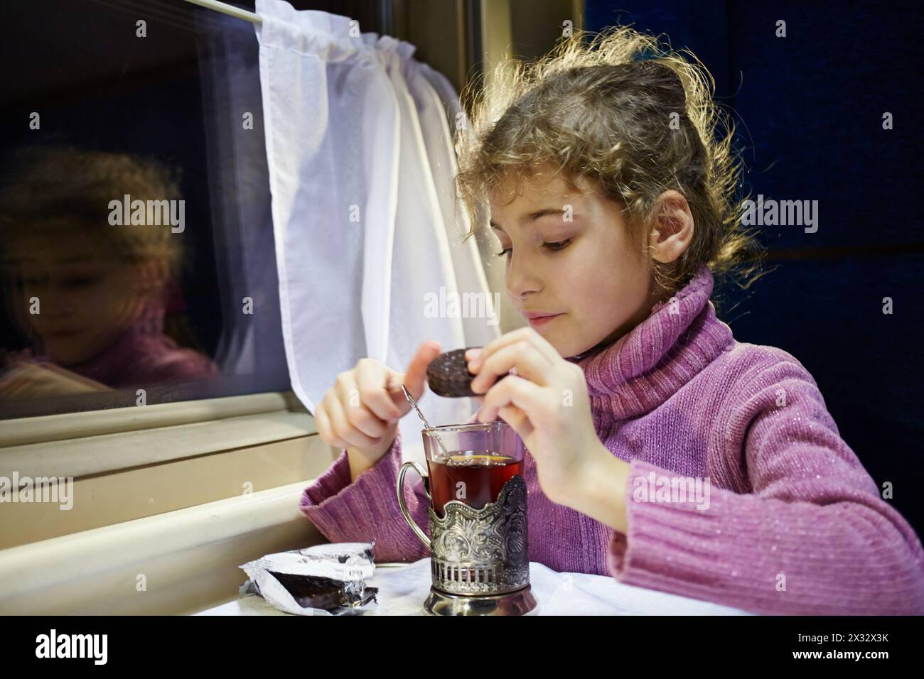Little girl drinks tea and eats chocolate cookie at table in coach ...