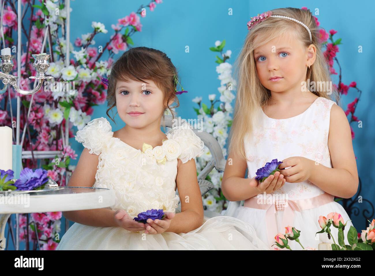 Two pretty little girls in white dresses hold blue flowers in room with ...