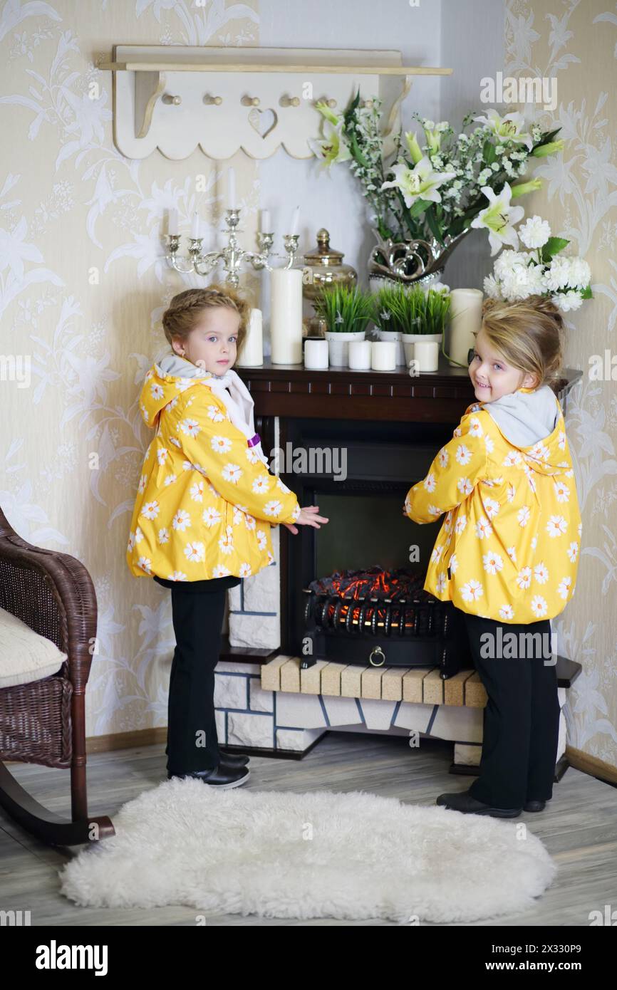 Two little girls in same clothes stands near fireplace and wicker ...
