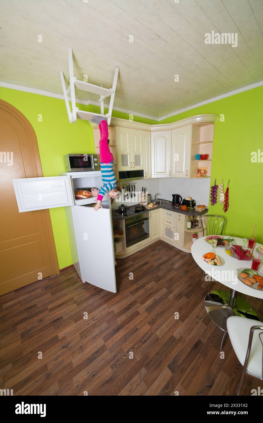 Little girl upside down holds a fridge in the kitchen with table and ...