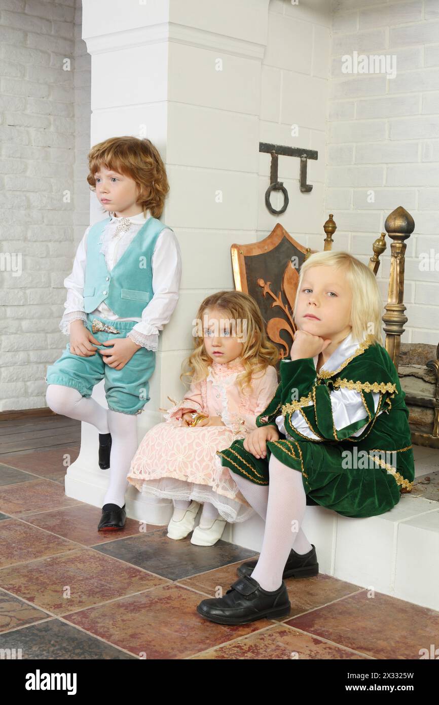 Two boys and girl in medieval costumes sit near fireplace with logs ...