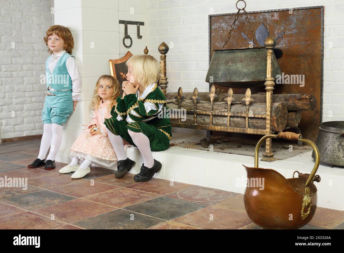 Two boys and girl in medieval costumes sit near fireplace with logs and ...