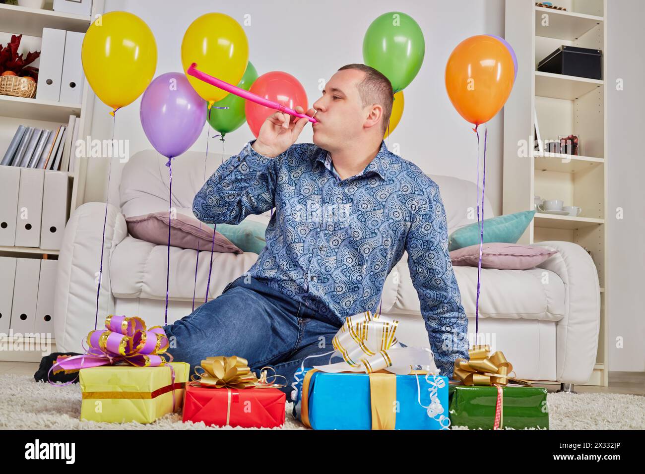 Man sits on carpet among gift boxes and birthday air balloons and blows ...