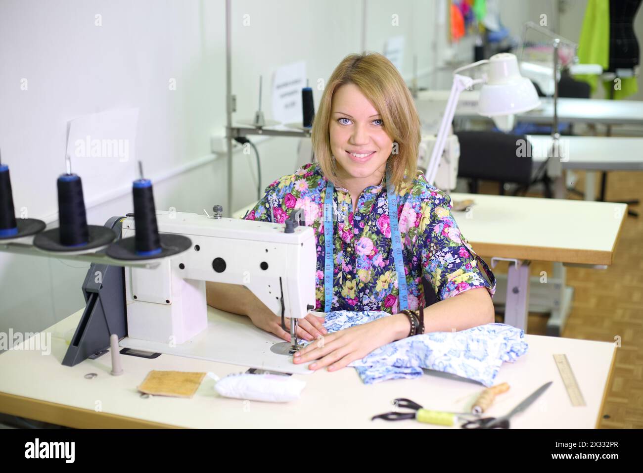 Pretty female tailor sits at table with sewing machine, sews and looks ...