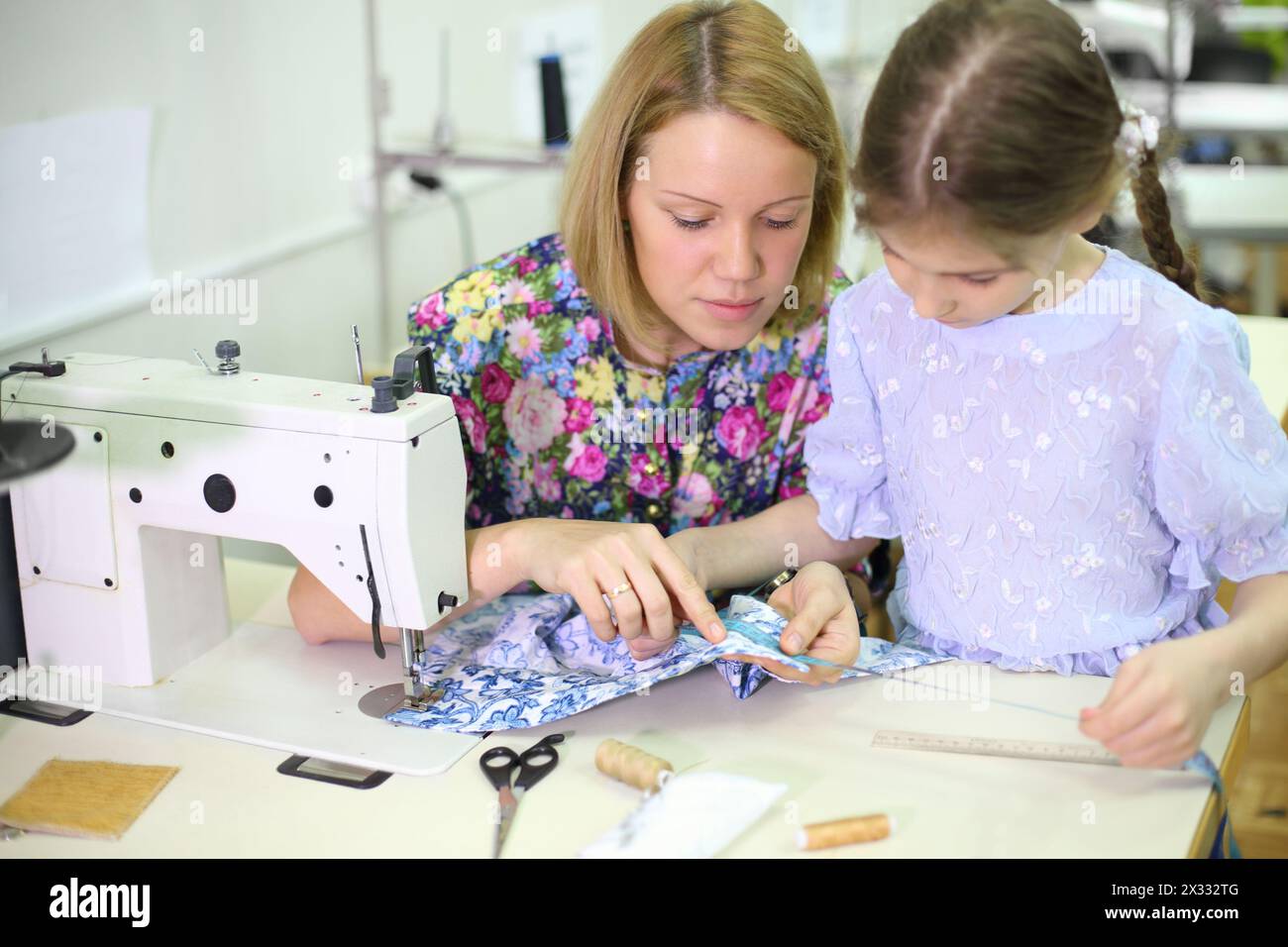 Female tailor sits at table with sewing machine and teaches girl how ...