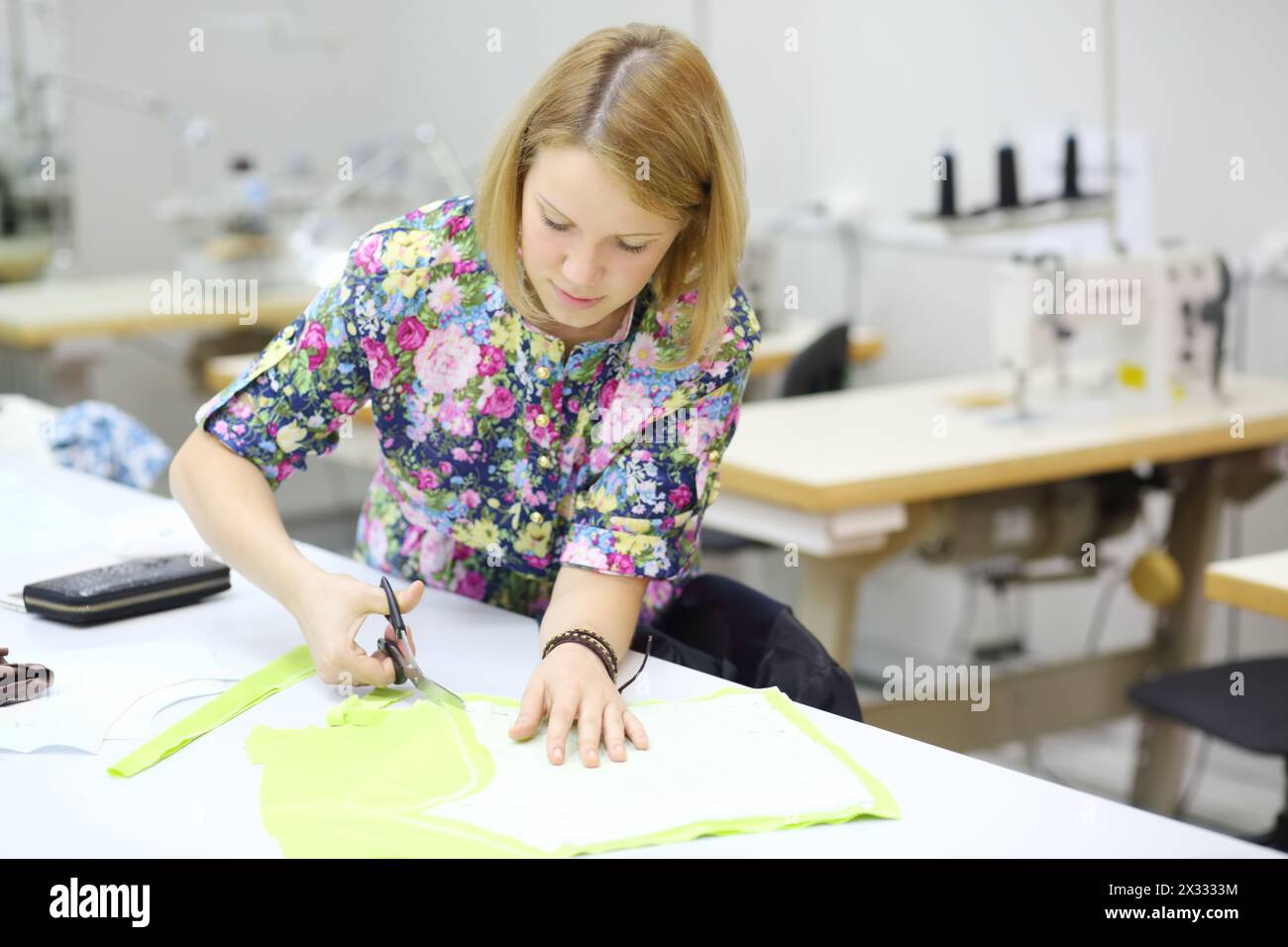 Female tailor stands near table and cuts tailoring detail at green ...