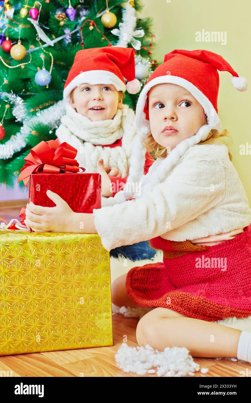 Little boy and girl in santa caps sit on furry rug under christmas tree ...
