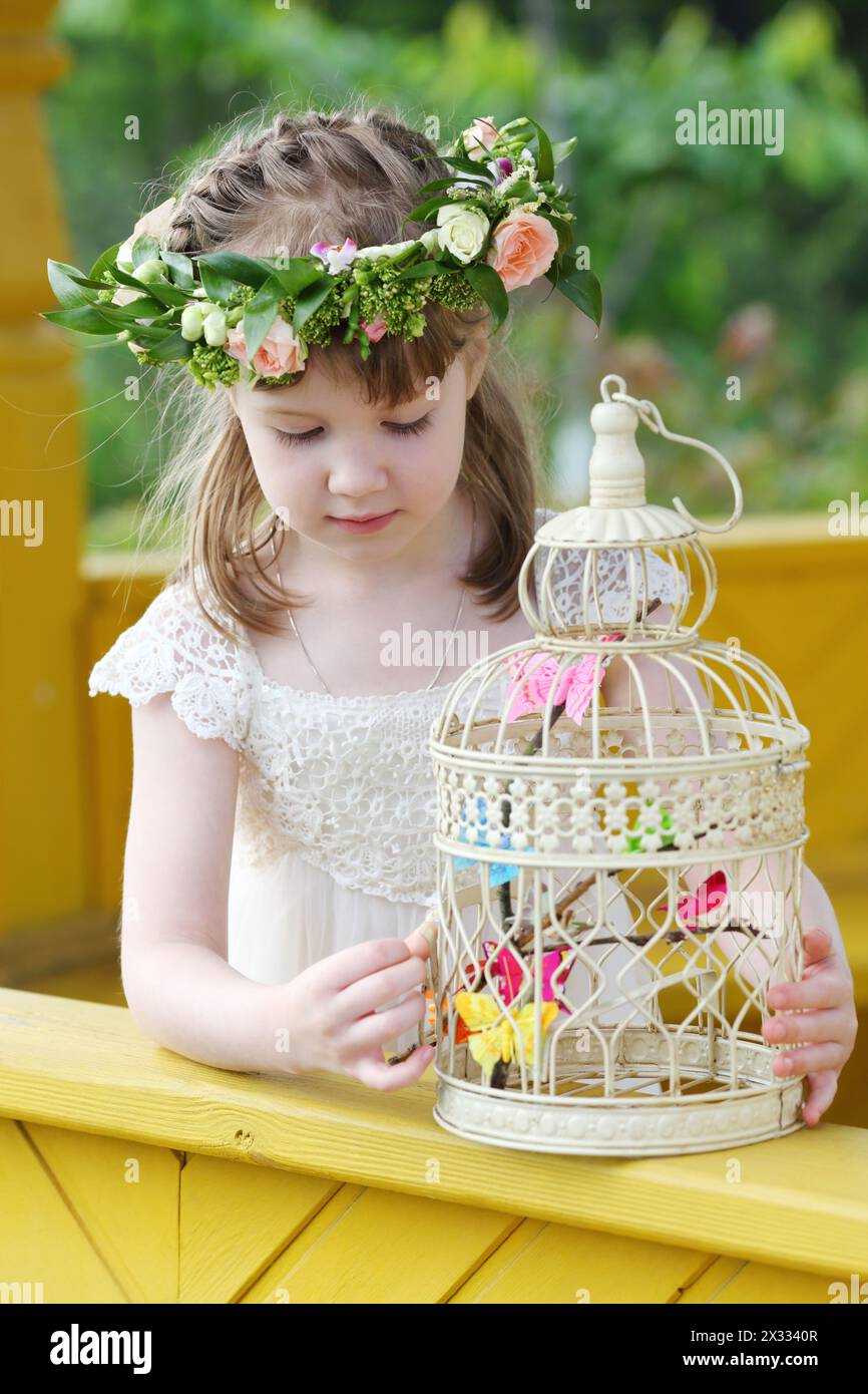 Little pretty girl in white dress and wreath plays with cage with ...