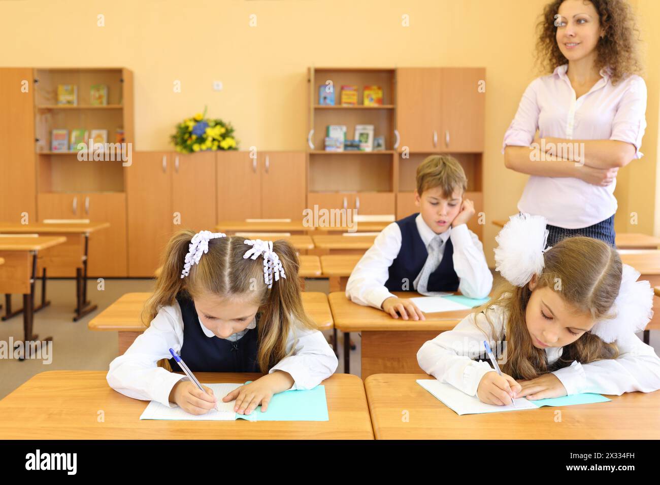 Two girls and boy write at school desks in classroom and teacher smiles ...