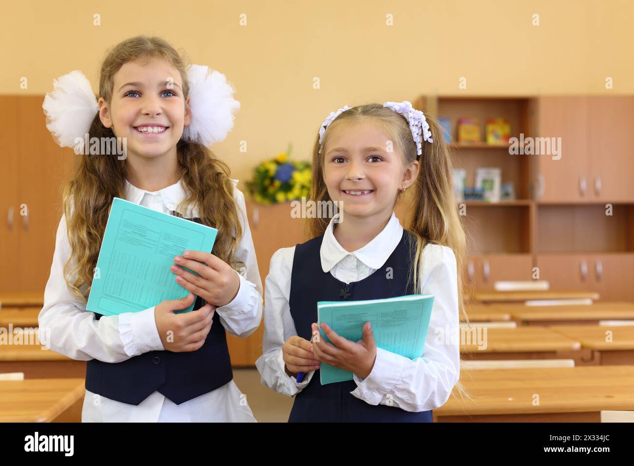 Two happy girls in uniform stand in classroom and hold green exercise ...