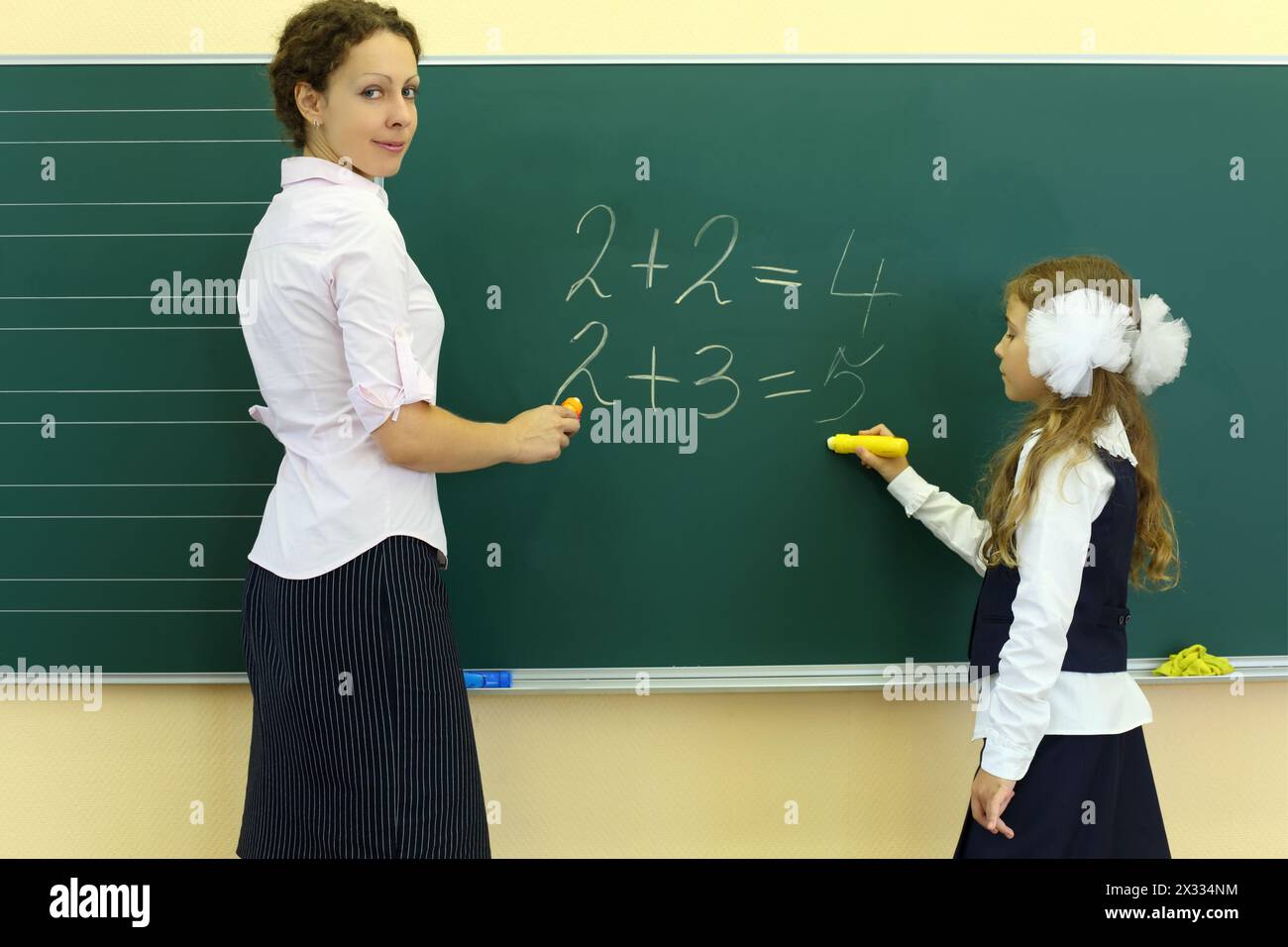 Girl and teacher stand near chalkboard and solve simple math examples ...