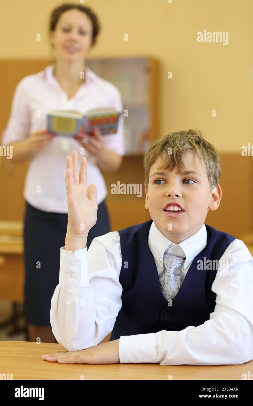 Teacher with book stands and boy sits at desk raised his hand to answer ...