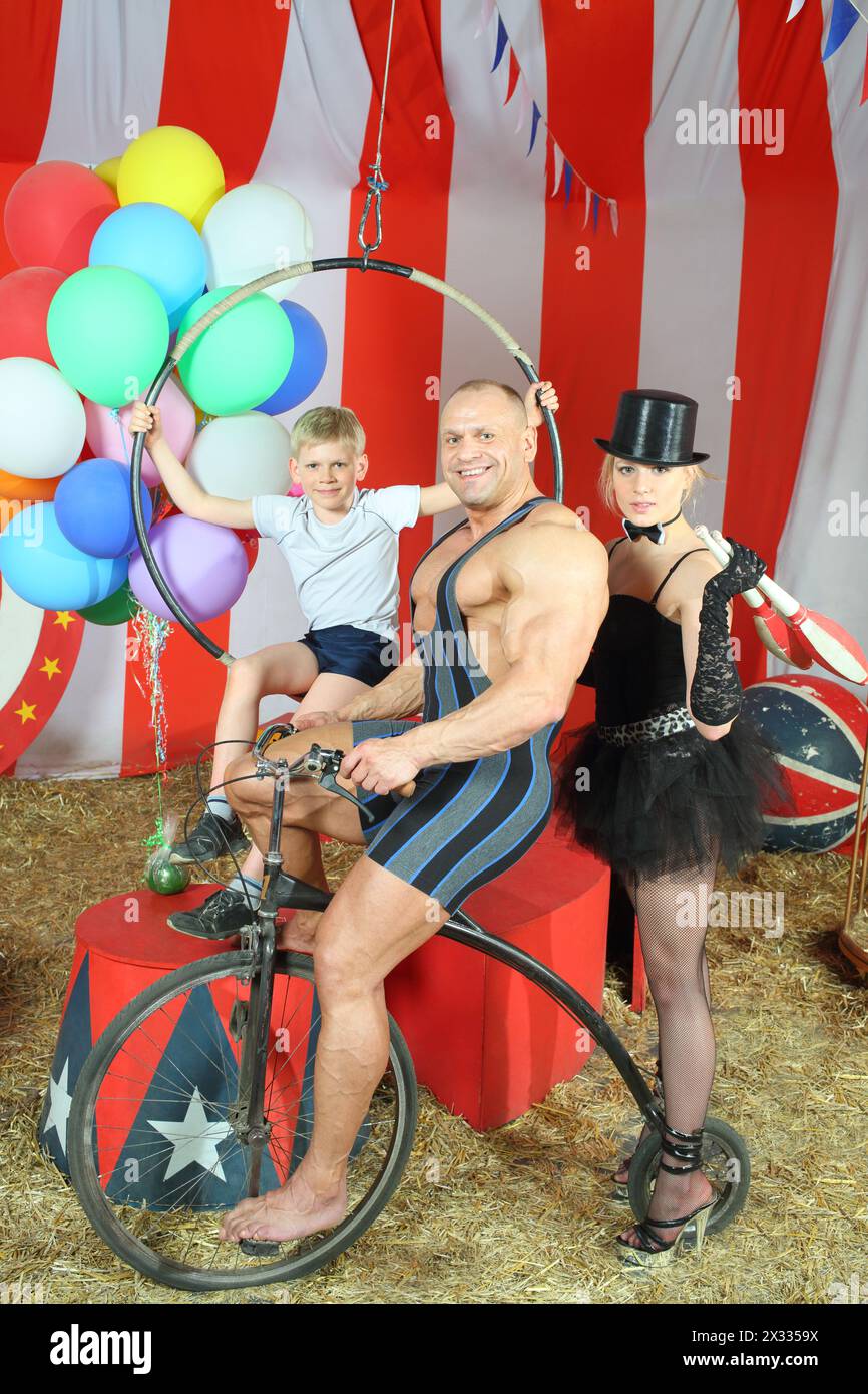 Three happy circus strong man, woman in cylinder and boy on retro bike with balloons in striped ...