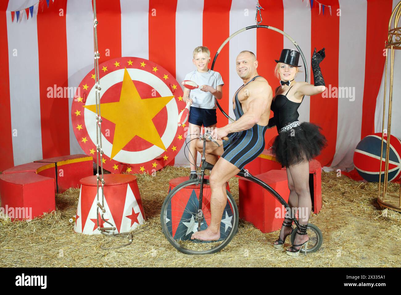 Three funny circus strong man, woman in cylinder and boy on retro bike with balloons in striped ...