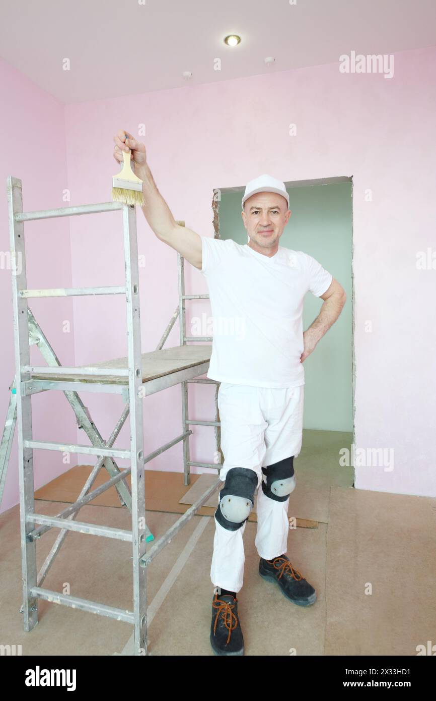 Happy plasterer in white clothes and kneepads standing near scaffolding ...