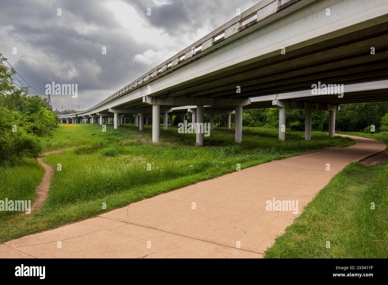 A paved pathway winds through a green area underneath a long, elevated ...