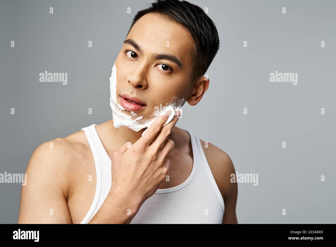 Handsome Asian man with shaving foam on face, meticulously shaving with a razor in a grey studio ...