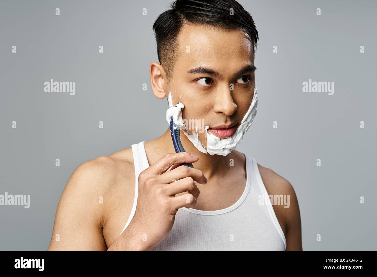 A portrait of a handsome Asian man with shaving foam on his face, carefully shaving with a razor ...