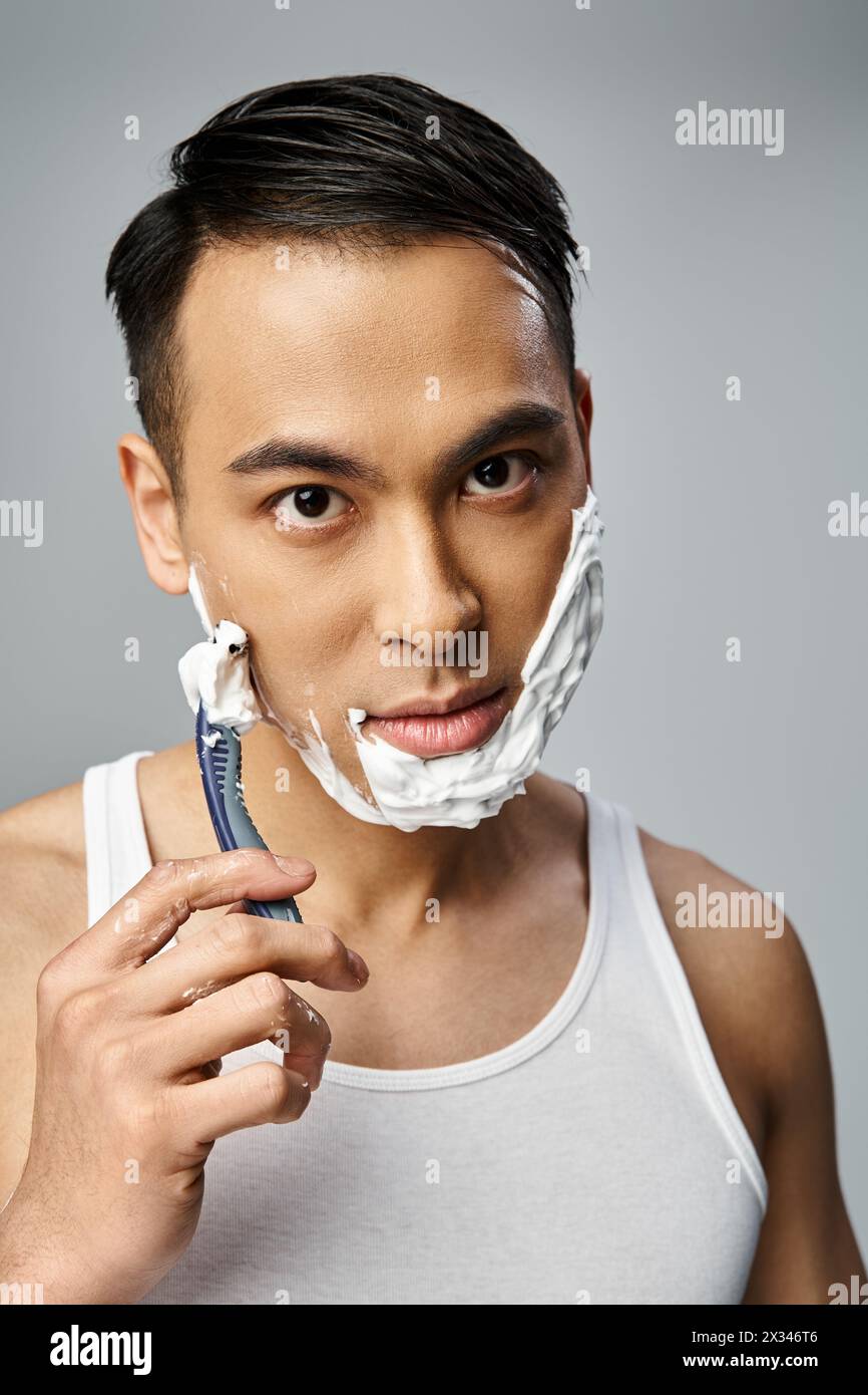 An Asian man with shaving foam on his face attentively shaves with a razor in a serene grey ...