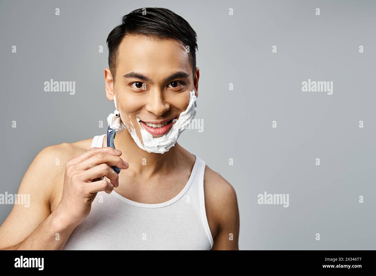 A handsome Asian man with shaving foam on his face is carefully shaving with a razor in a grey ...
