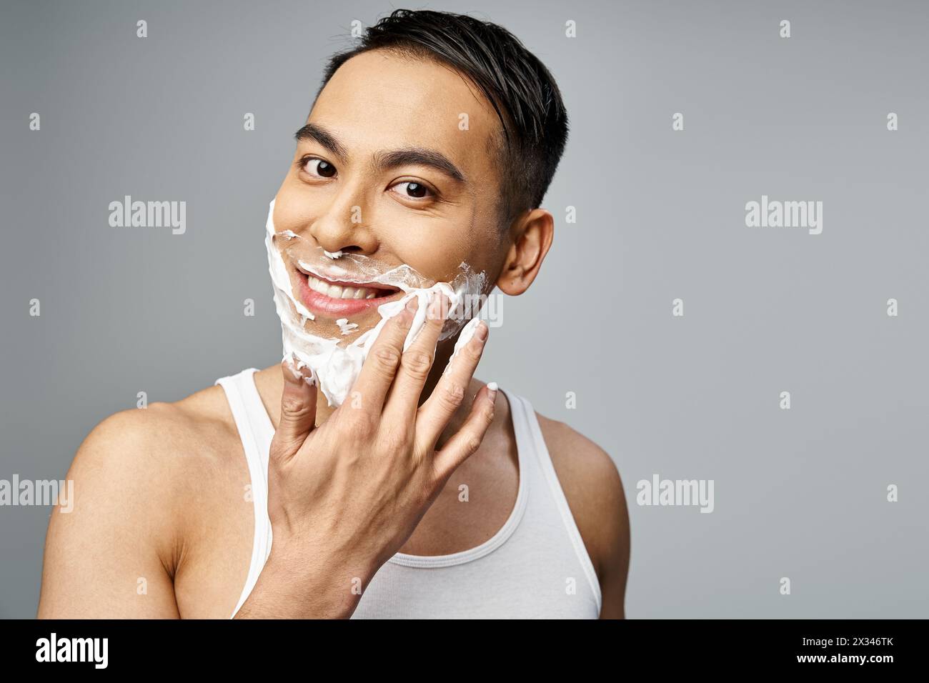 A handsome Asian man with shaving foam on face, meticulously shaving with a razor in a grey ...