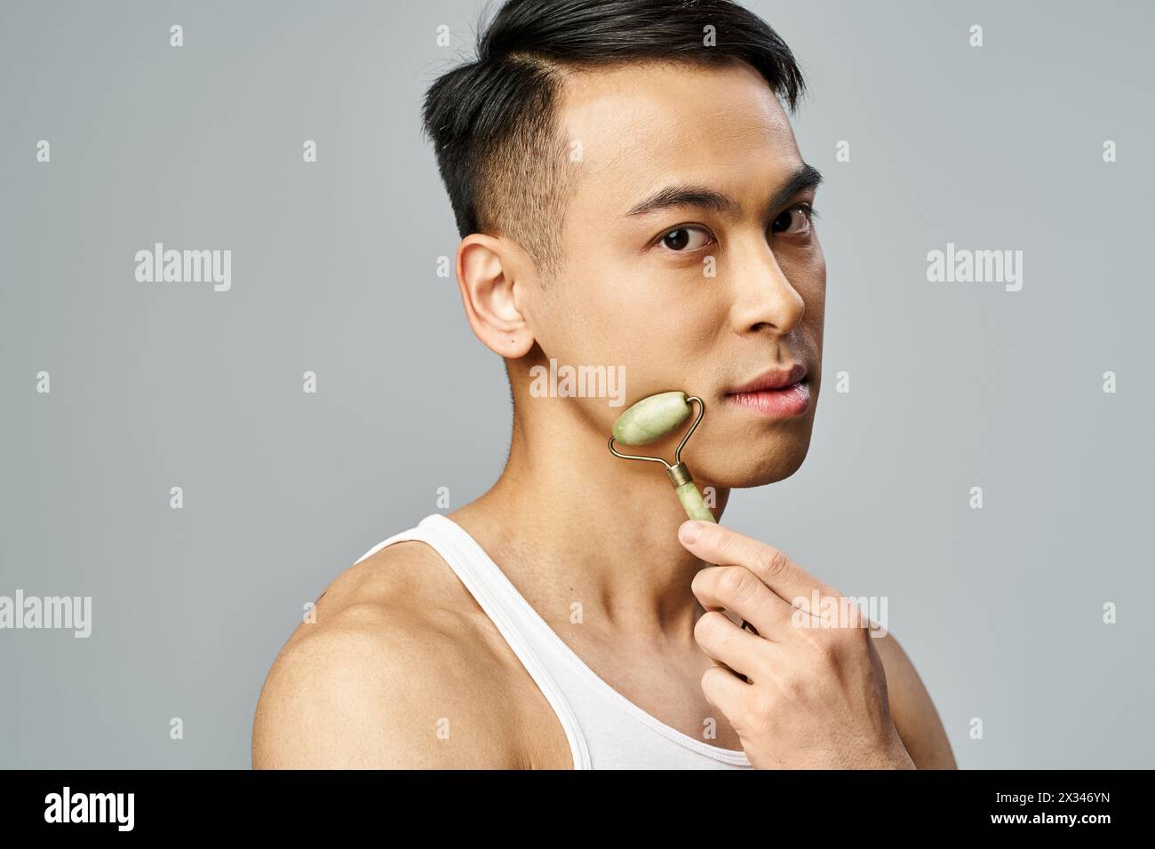 Asian man using jade roller, focusing on grooming and self-care routine in a grey studio setting ...