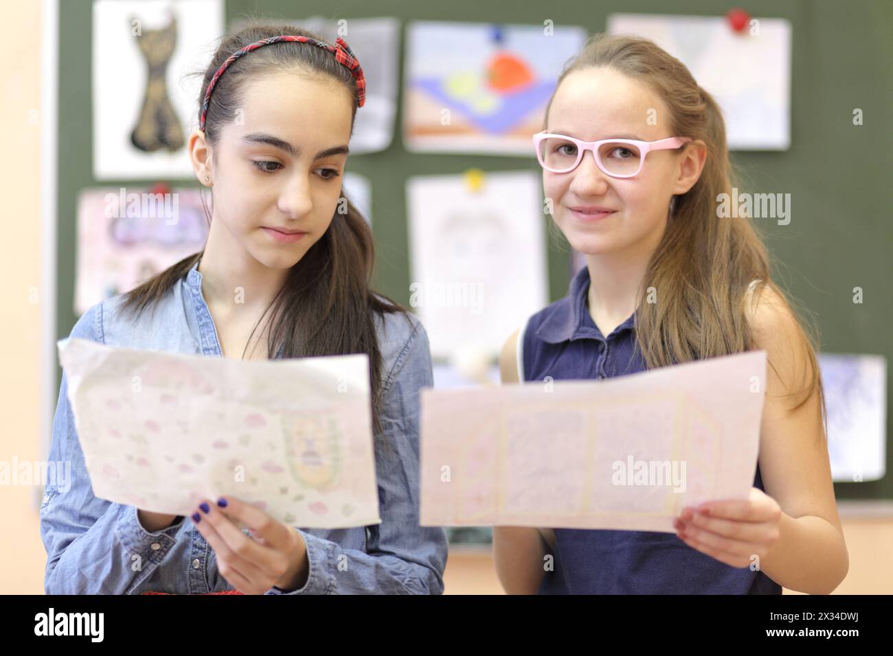 Two schoolgirls consider drawings on drawing lesson in classroom, one ...
