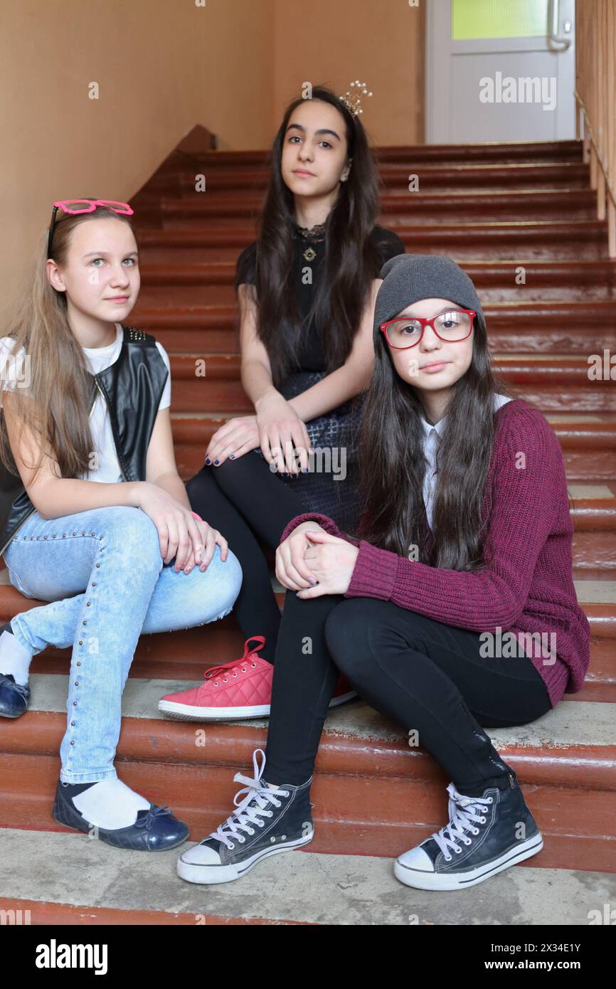 Three girls sitting together on stairs at school, looking into camera ...