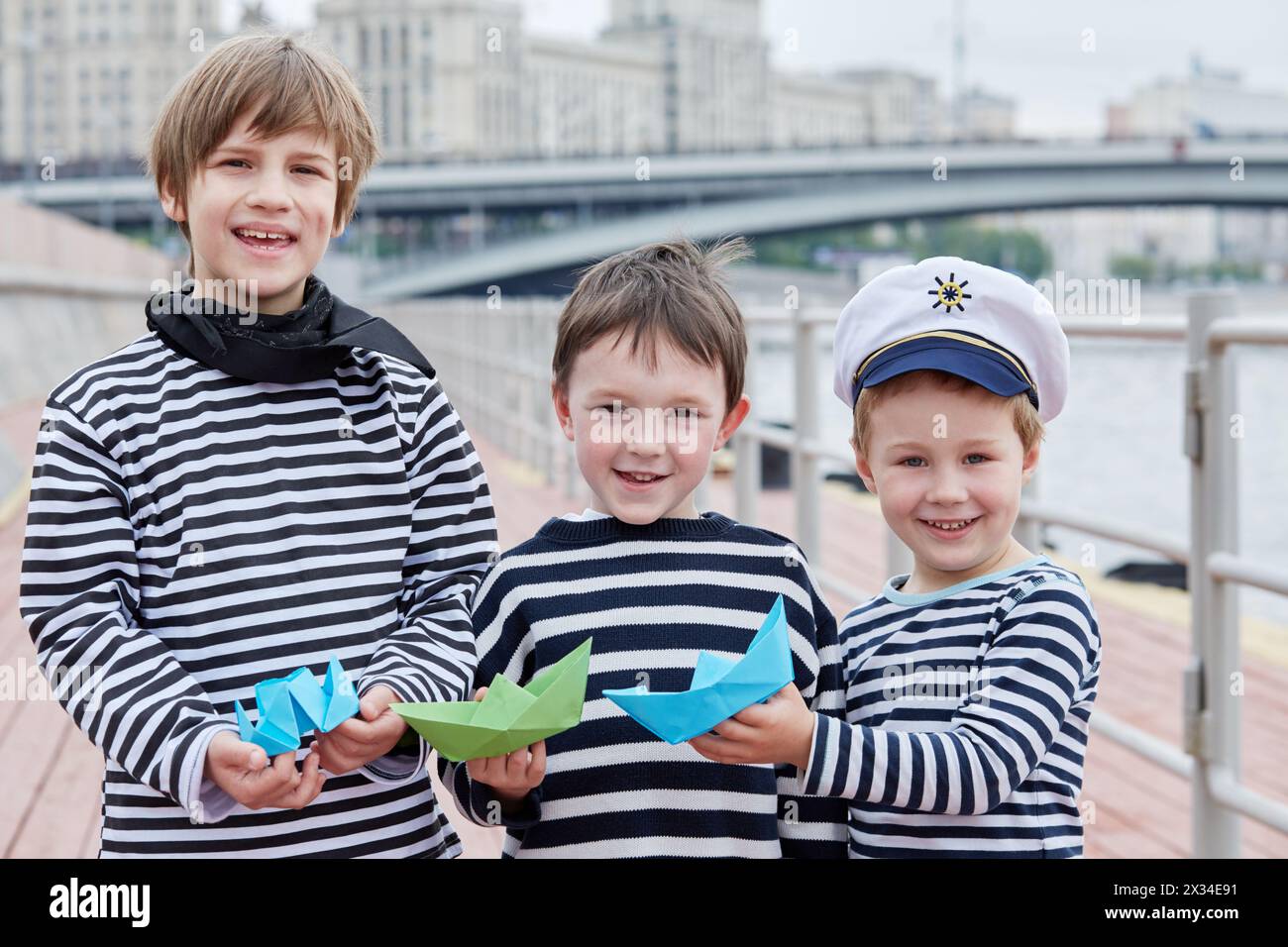 Three smiling boys in striped vests play with paper boats on embankment ...