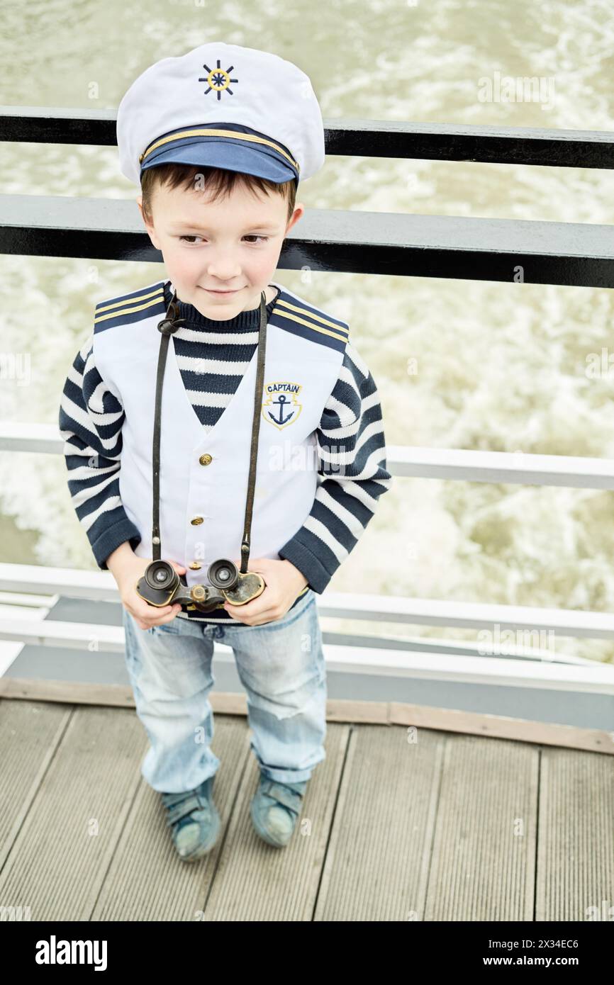 Little boy dressed as a captain stands with binoculars at boat stern ...